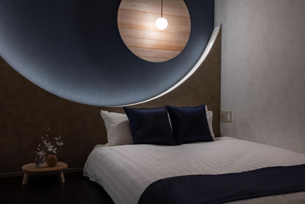 Quarto moderno com luminária circular no teto, cama com roupa de cama branca e uma pequena mesa redonda com um vaso.