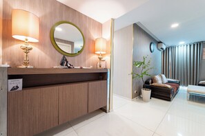 Um hall de hotel moderno com balcão de recepção de madeira, duas luminárias fixadas na parede, um espelho redondo, um relógio e um espaço de estar com sofá e poltronas.