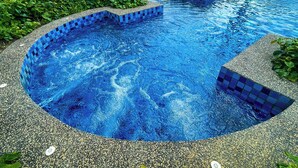 Piscina com borda de mosaico azul, rodeada por vegetação e um deck de pedriscos.