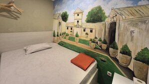 Quarto com um mural de uma cena de aldeia, uma cama com colchão branco e manta laranja, e um cabide de madeira na parede.