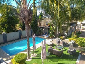 Piscina cercada por espreguiçadeiras e uma pequena área de jardim com palmeiras e arbustos.