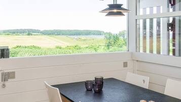 Quarto com mesa, cadeiras, vista para paisagem verde e luminária pendente.