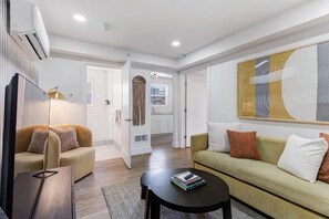 Uma sala de estar moderna com sofá, mesa de centro e televisão. Há uma porta que dá para outro quarto.