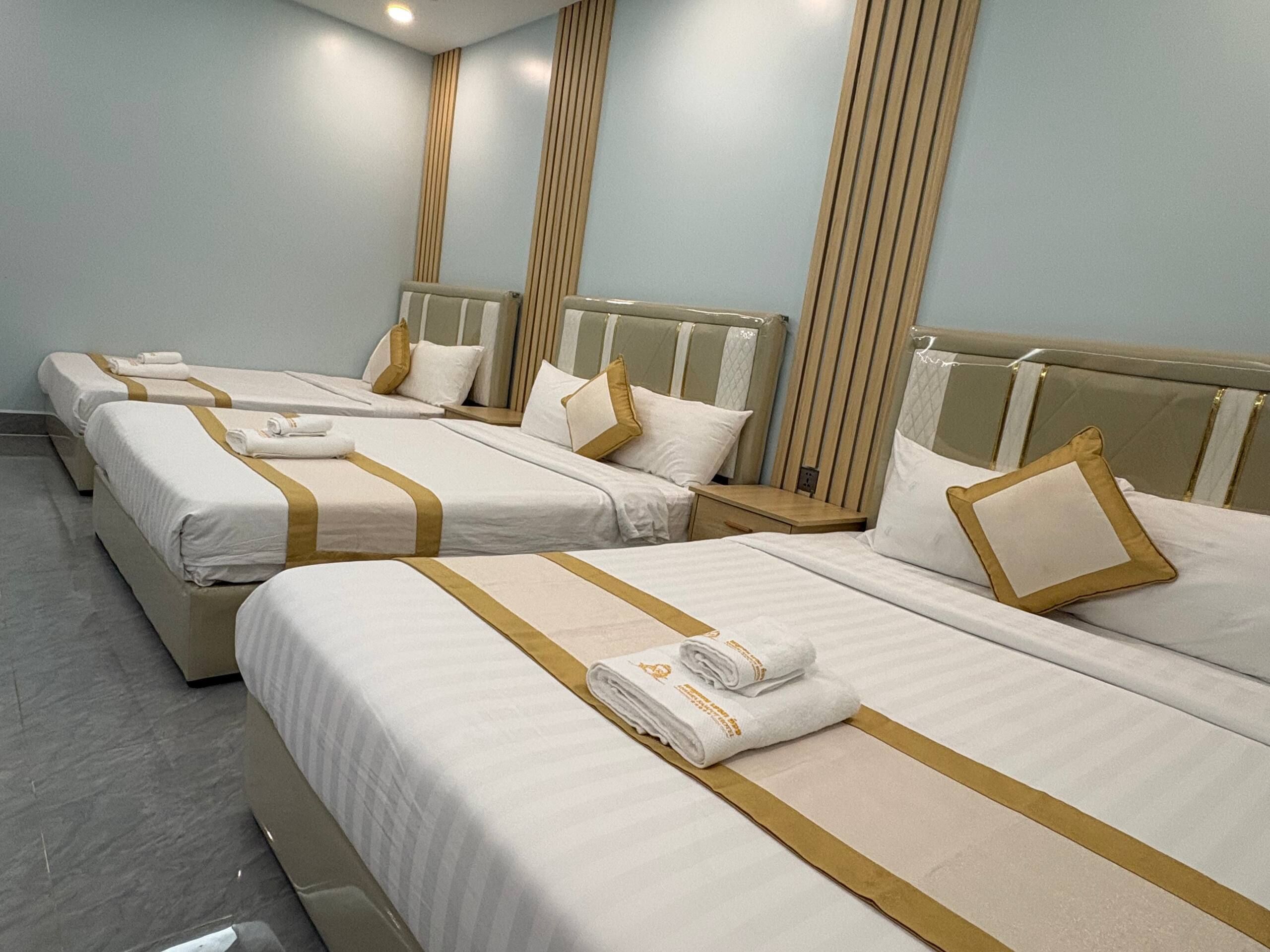 Quarto de hotel com quatro camas, cada uma com roupa de cama branca e detalhes dourados, e cabeceira de madeira.