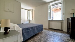 Quarto com cama, radiador, janela com cortinas, escrivaninha com luminária e cômoda.