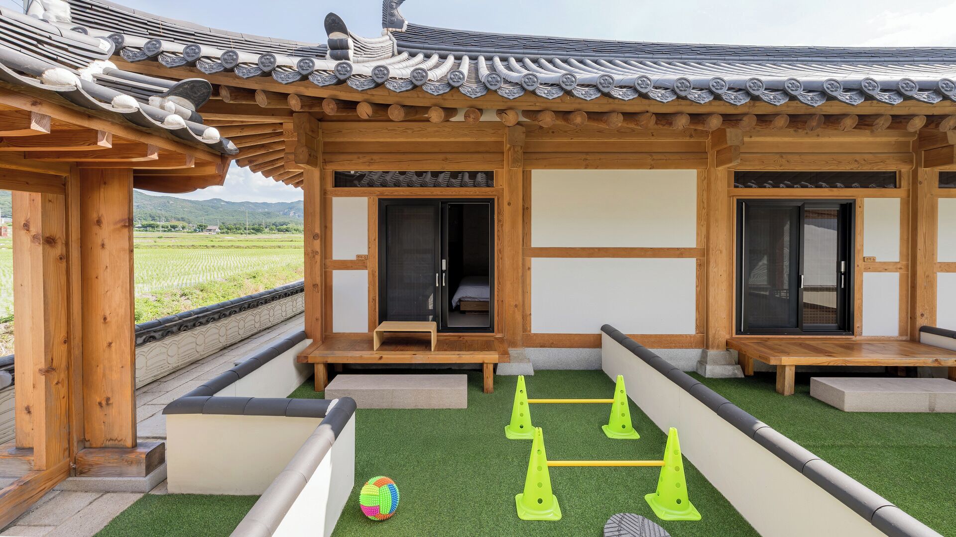 Um edifício tradicional em estilo japonês, com arquitetura de madeira, vista para um campo verde e uma pequena área de recreação com cones e uma bola.