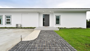 Um edifício moderno com teto plano, entrada central com porta escura e entrada de automóveis pavimentada que leva à entrada.