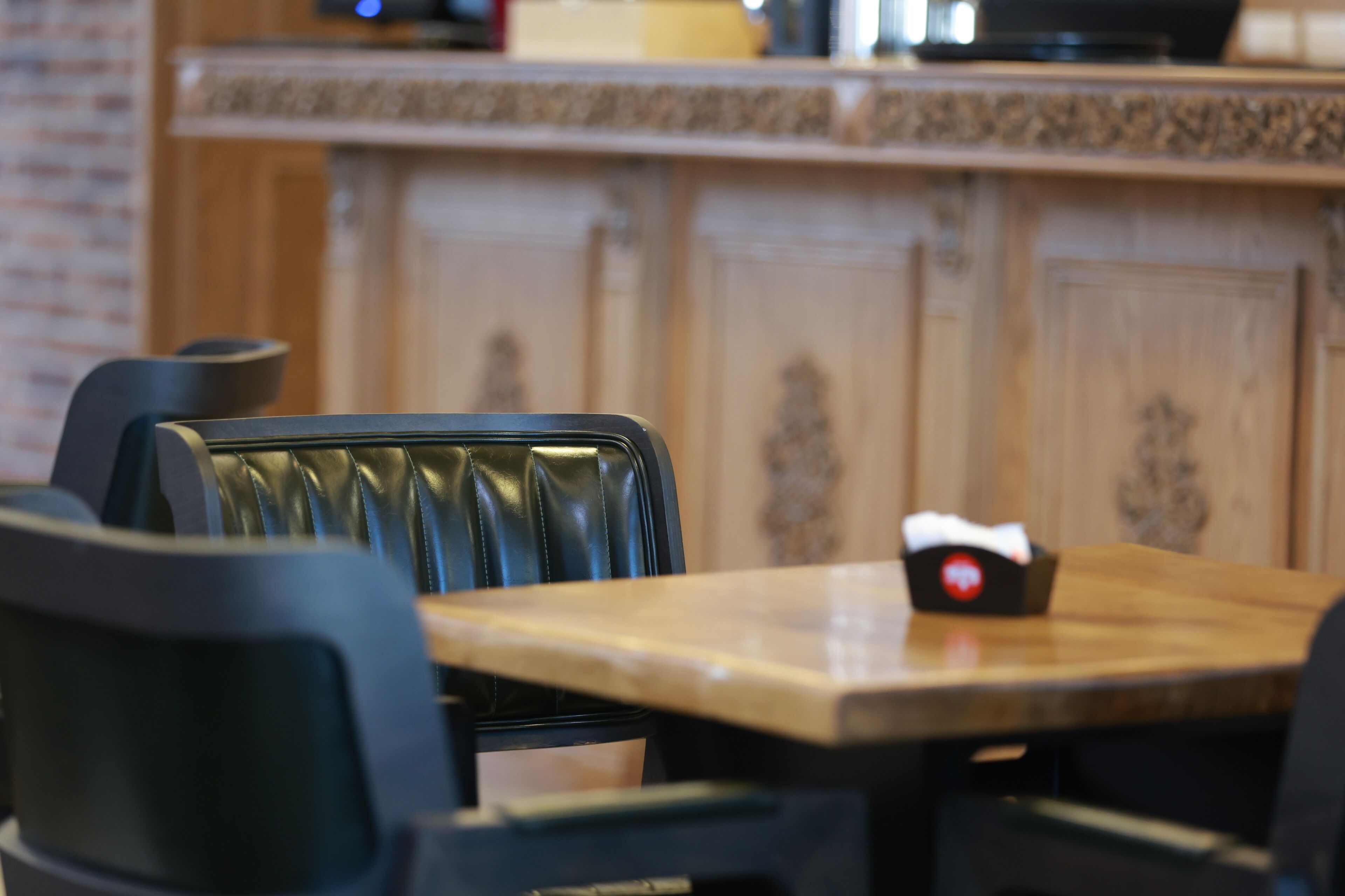 Interior de restaurante com mesa de madeira, cadeiras de couro preto e balcão com logotipo vermelho.