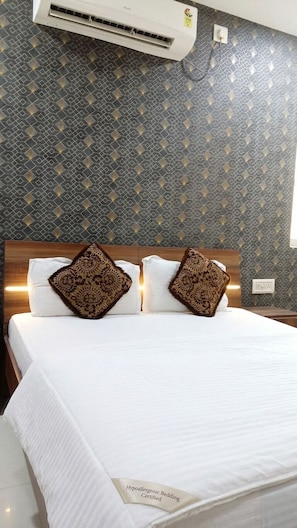 Quarto de hotel com uma cama, duas almofadas estampadas, um protetor de colchão branco e um ar condicionado fixado na parede.