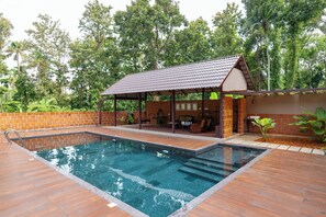 Piscina com deck de madeira, área de estar coberta e muro de tijolos.