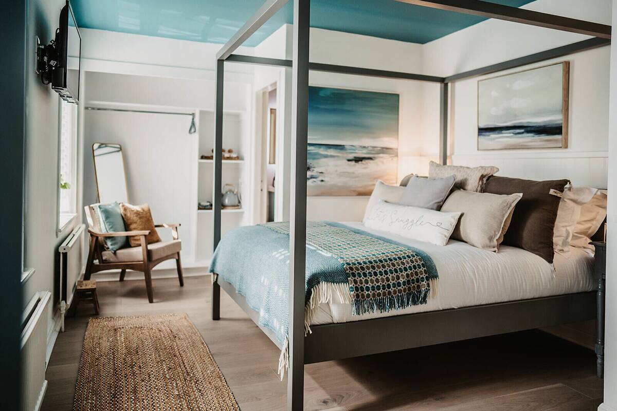 Uma cama de dossel com vista para o mar, uma poltrona com almofadas, um espelho e uma prateleira com objetos.