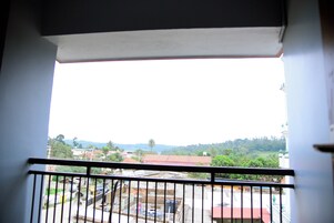 Uma varanda com grades de metal, vista para uma rua com prédios e paisagem distante com árvores e colinas.