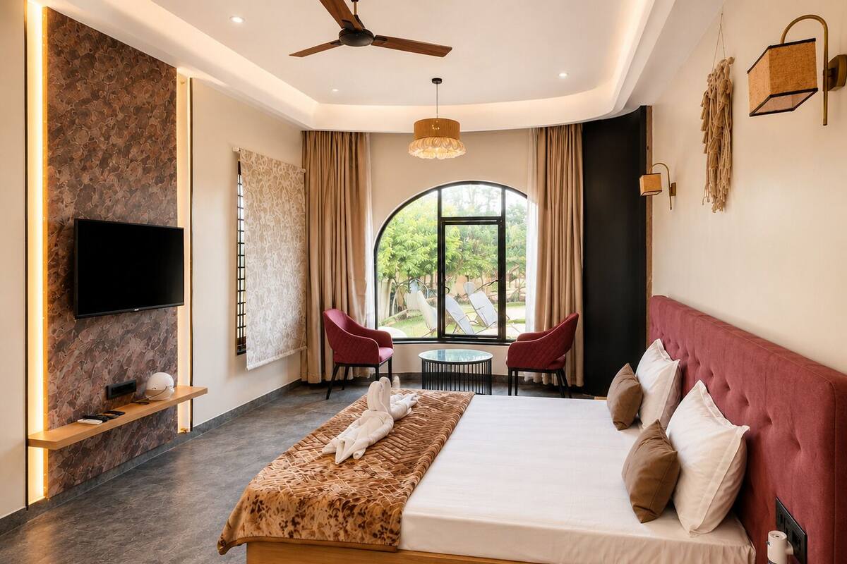 Quarto de hotel moderno com uma cama grande, ventilador de teto, televisão, janela com vista para vegetação e uma pequena mesa com cadeiras.