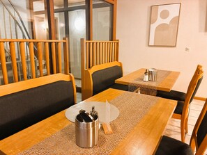Uma área de refeições com mesas e cadeiras de madeira, uma cafeteira de metal e um jogo de mesa estampado.