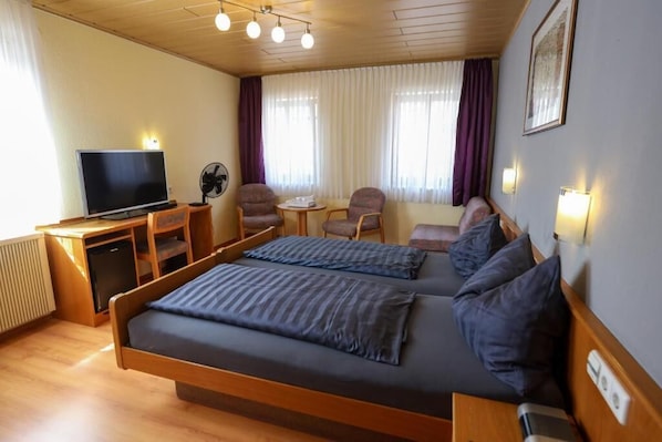 Quarto de hotel com cama, escrivaninha, televisão, ventilador e janela com cortinas.