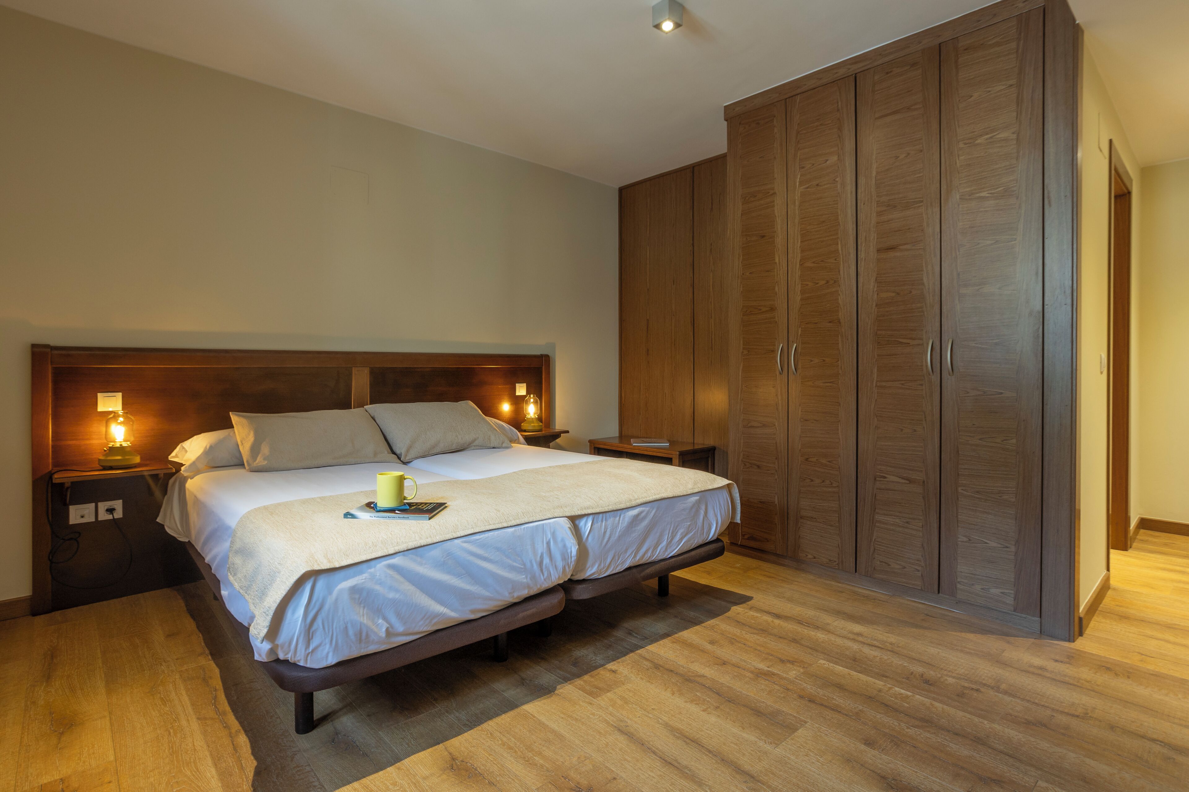 Quarto com cama grande, guarda-roupa de madeira, mesas de cabeceira com luminárias e um criado-mudo.