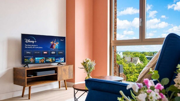 Uma sala de estar com uma televisão exibindo conteúdo do Disney+, uma poltrona azul, uma mesinha e vista para o verde lá fora.