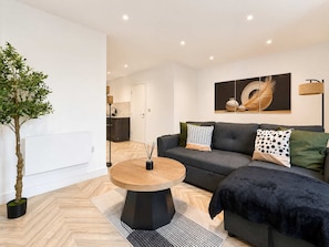 Uma sala de estar moderna com uma mesa de centro redonda de madeira, um sofá cinza com almofadas estampadas, um quadro abstrato grande e um vaso de planta.