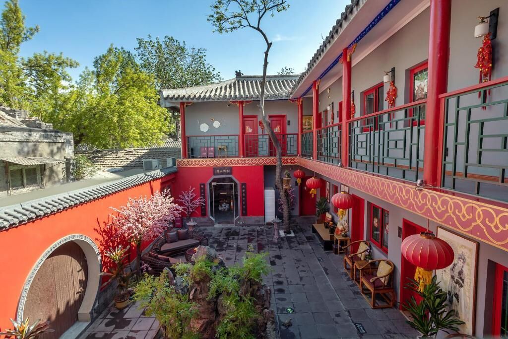 Pátio com arquitetura tradicional, incluindo um edifício vermelho com telhado de azulejos, um caminho de pedra e uma área de estar com cadeiras.