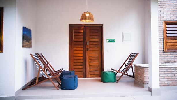Uma porta de madeira, duas cadeiras listradas, um lustre pendurado e uma bolsa verde em um alpendre.