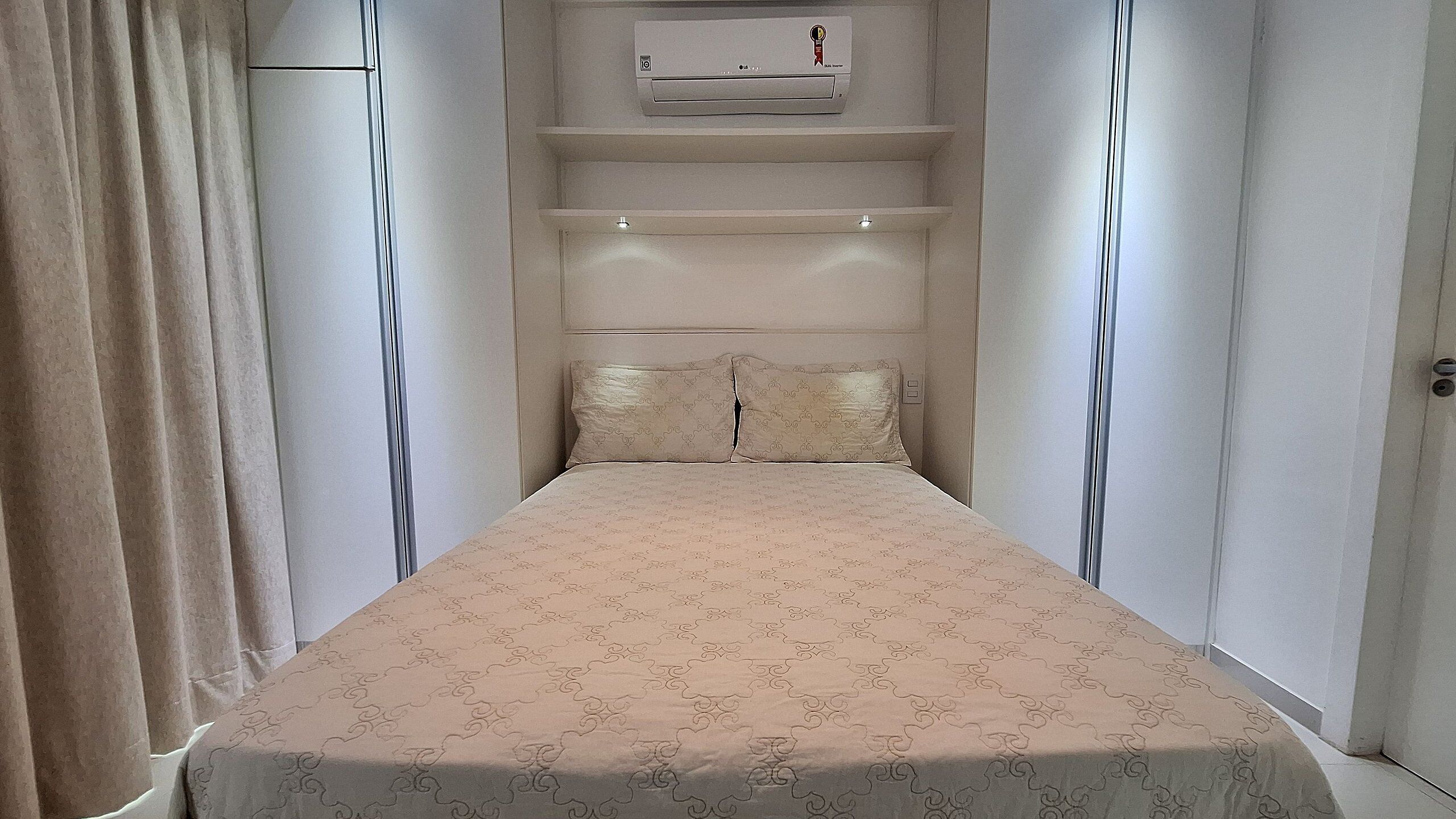 Quarto pequeno com cama, ar condicionado no teto e uma porta de um lado.