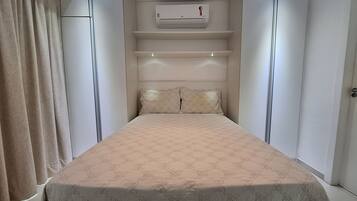 Quarto pequeno com cama, ar condicionado no teto e uma porta de um lado.
