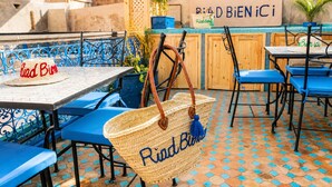 Uma área de estar ao ar livre com cadeiras e mesas azuis, uma cesta de vime com o texto 'Riad BieniCi' e uma placa com a inscrição 'Riad BieniCi'.