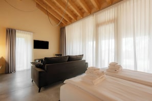 Quarto de hotel com cama, sofá, televisão de tela plana e janelas amplas com cortinas.