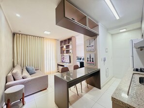 Quarto de hotel moderno com sofá, mesa de jantar de vidro, área de cozinha e vista para a paisagem urbana.