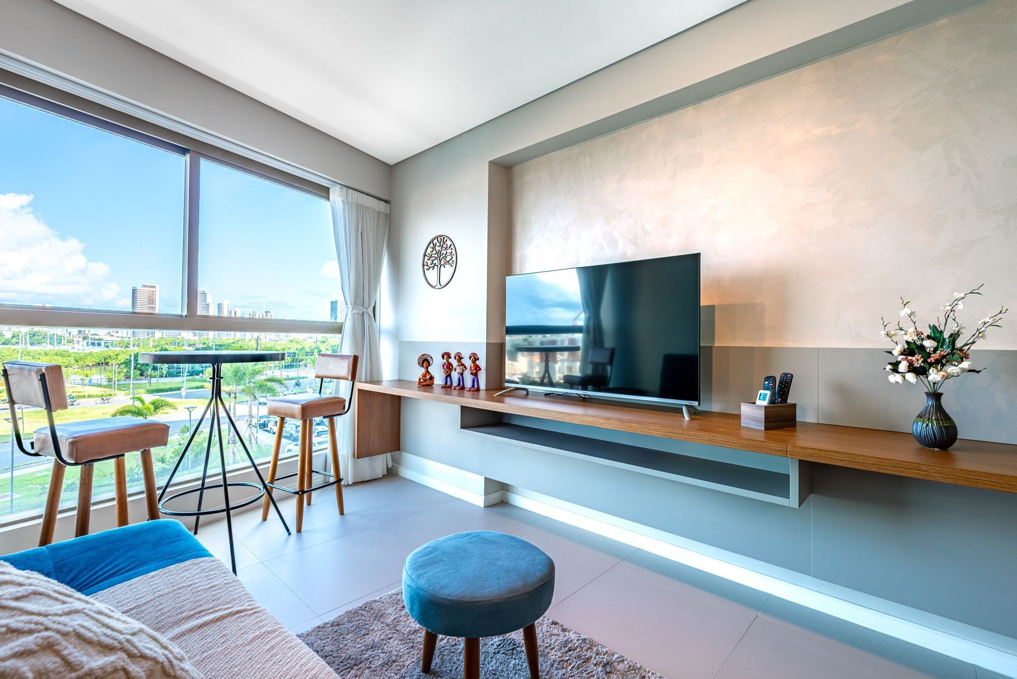 Sala moderna com TV grande, área de jantar com cadeiras, sofá e vista panorâmica da cidade através de amplas janelas.