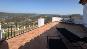 Terraço com vista para um lago e colinas, mobiliado com mesa e cadeiras.
