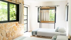 Quarto com duas camas, parede de pedra, janela com vista para o verde e um banco de madeira.