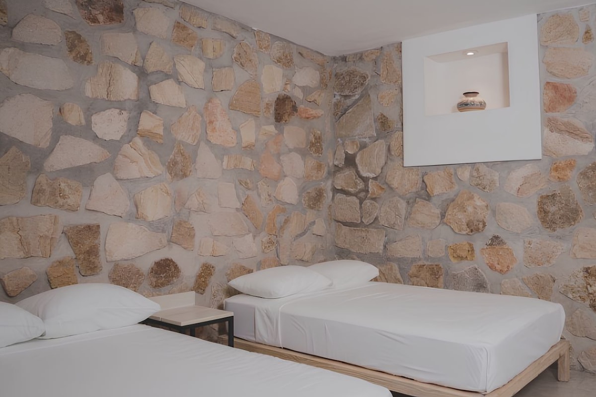 Quarto com parede de pedra, duas camas, uma mesa de cabeceira e um quadro pendurado na parede.