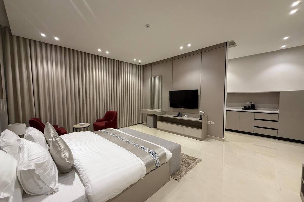 Quarto de hotel moderno com uma cama grande, televisão, área de estar com cadeiras vermelhas e uma cozinha compacta.