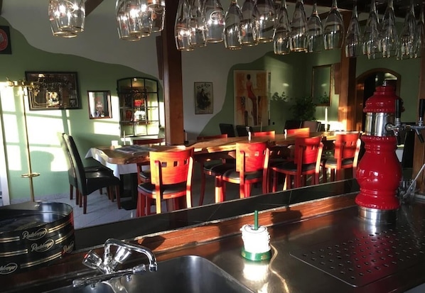 Interior de restaurante com área de bar, cadeiras vermelhas, máquina de café vermelha e luminárias pendentes.