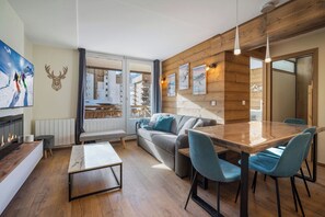 Uma sala de estar moderna com teto de madeira, lareira, sofá, mesa de jantar com cadeiras e vista para uma montanha através de uma porta de vidro.