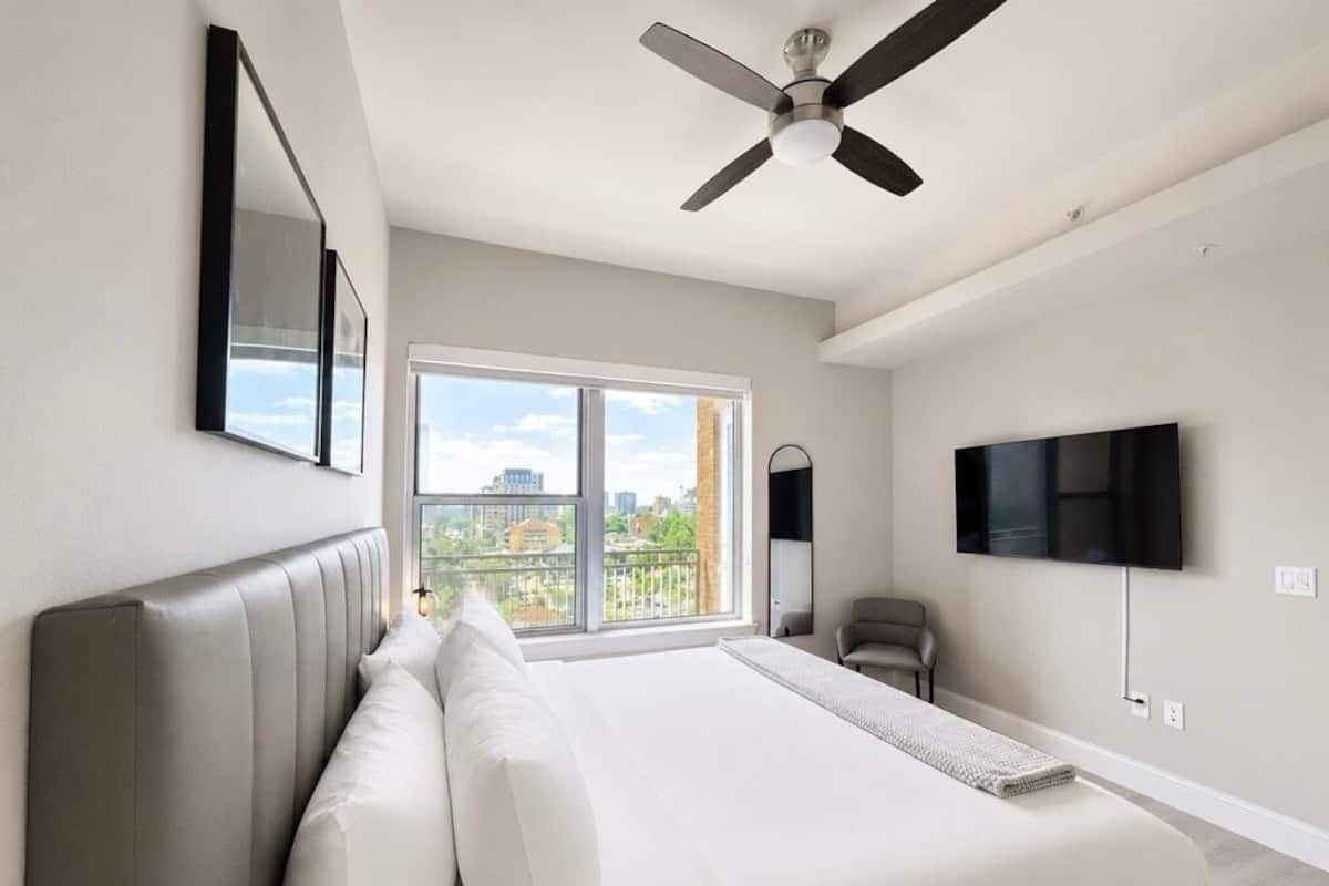 Quarto com cama grande, ventilador de teto, televisão e janela com vista para a cidade.