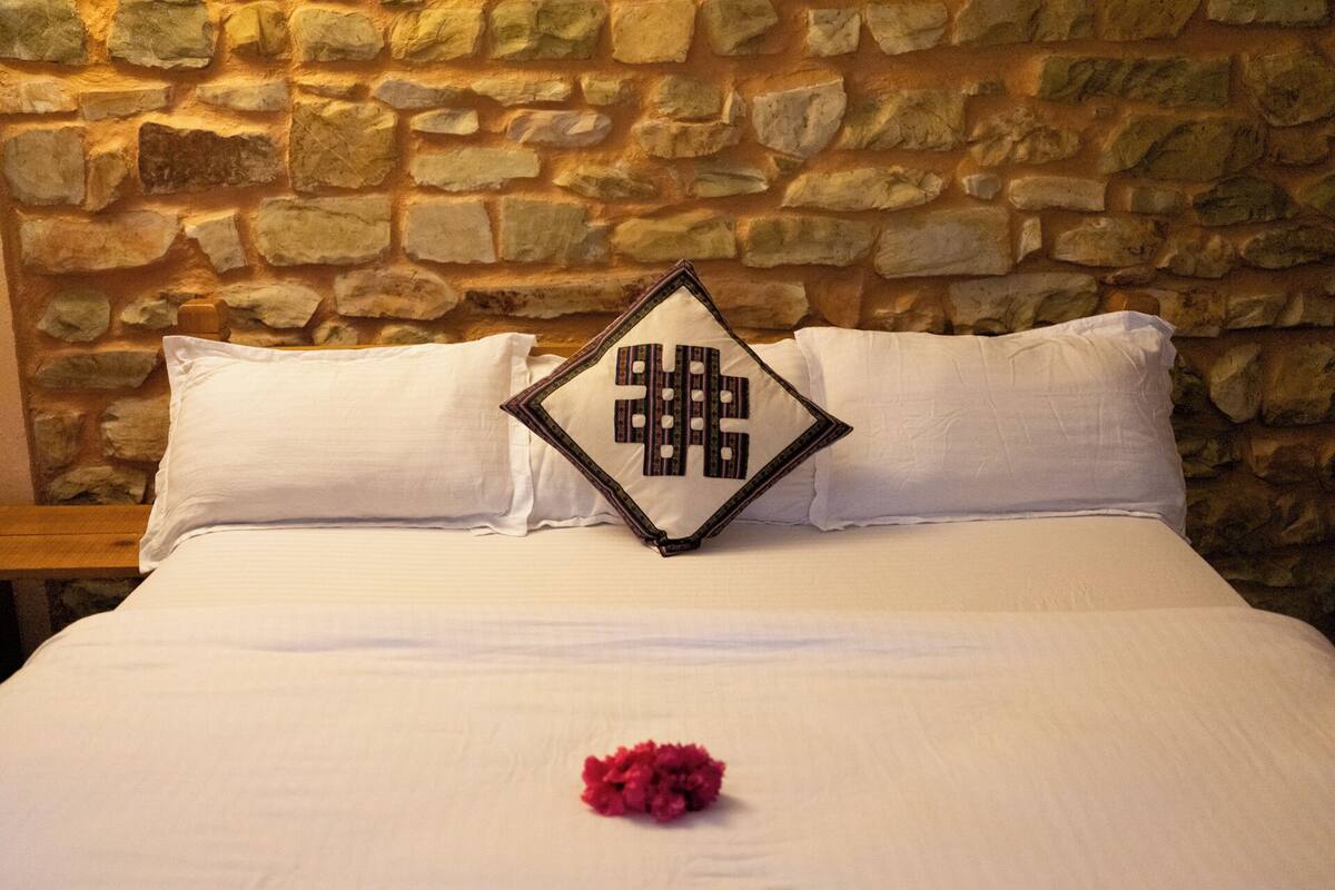 Uma cama com roupa de cama branca, um travesseiro decorativo e uma parede de pedra ao fundo.