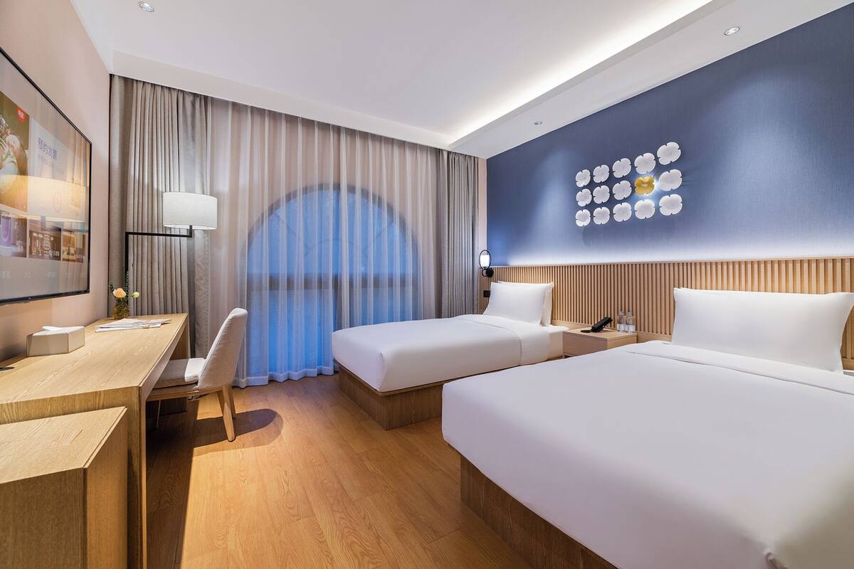 Quarto de hotel com duas camas, uma escrivaninha, uma cadeira, um abajur e uma parede com padrão floral.