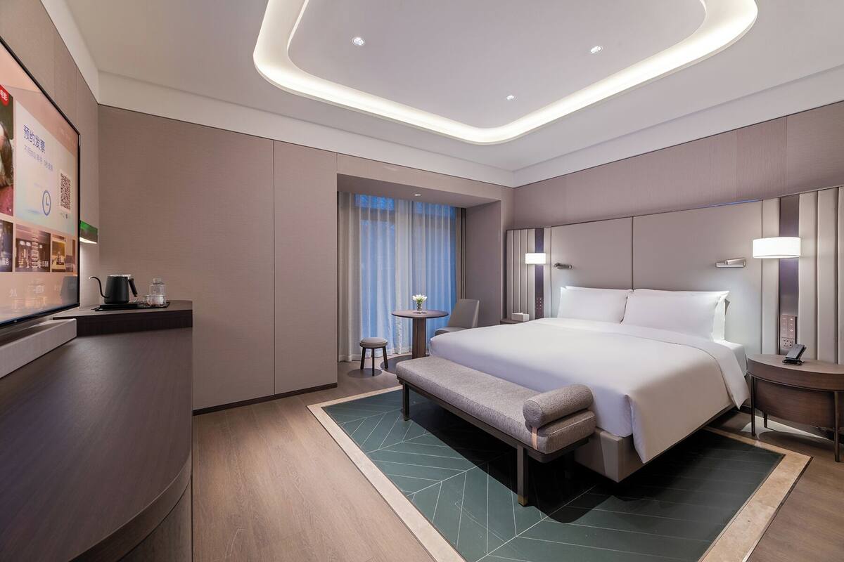 Quarto de hotel moderno com uma cama grande, uma mesa com chaleira, uma mesinha com um vaso, e um espelho amplo.
