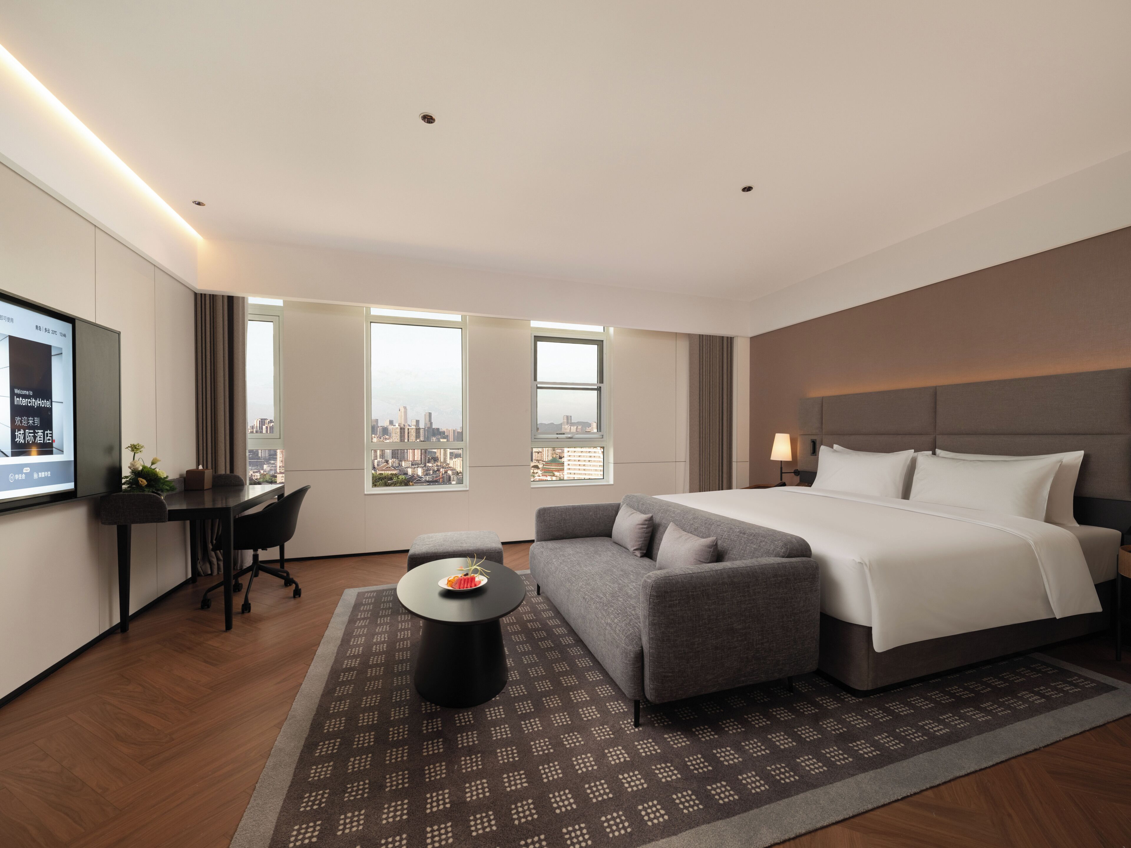 Quarto de hotel moderno com uma cama grande, um sofá, uma escrivaninha e uma televisão exibindo uma mensagem de boas-vindas.