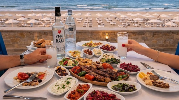 Mesa posta com diversos pratos e bebidas, com vista para uma praia com guarda-sóis.