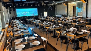 Um restaurante com várias mesas e cadeiras, uma tela grande exibindo uma imagem e uma escada que leva a um andar superior.