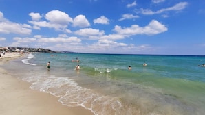 Uma praia com águas cristalinas, pessoas nadando e caminhando pela orla, e uma linha de costa distante com edifícios.