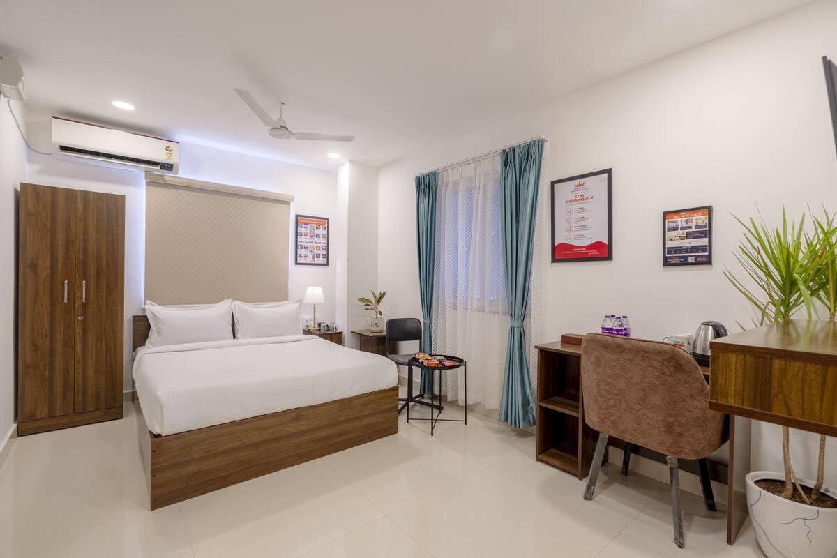 Quarto de hotel com cama, escrivaninha, cadeira, ar condicionado e uma planta em vaso.