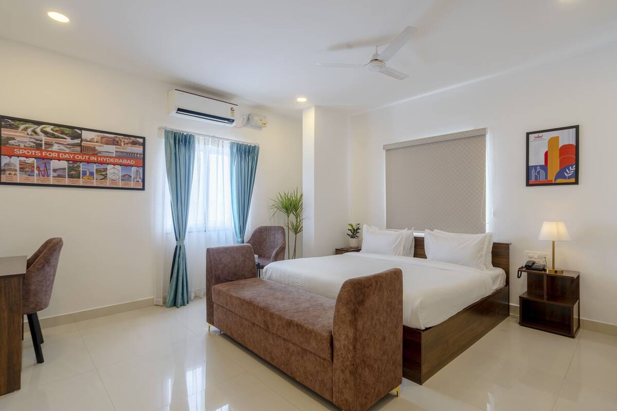 Quarto de hotel com cama, sofá, escrivaninha, cadeira, mesa de cabeceira, luminária, ar-condicionado, ventilador de teto, janela com cortinas e um pôster na parede.