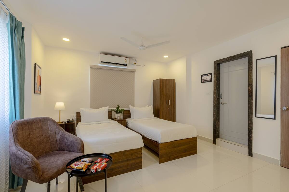 Quarto de hotel com duas camas, uma cadeira, uma mesa com lanches, ar condicionado, ventilador de teto e uma porta.