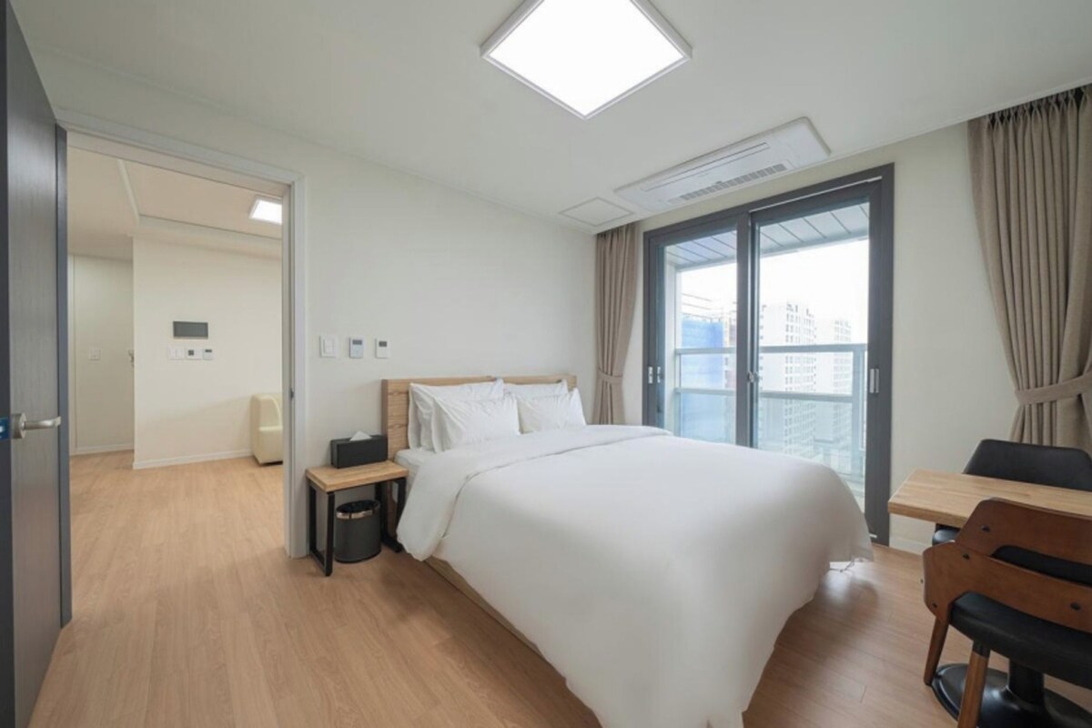 Quarto de hotel com uma cama grande, uma escrivaninha, uma cadeira, janela com cortinas e vista para edifícios.