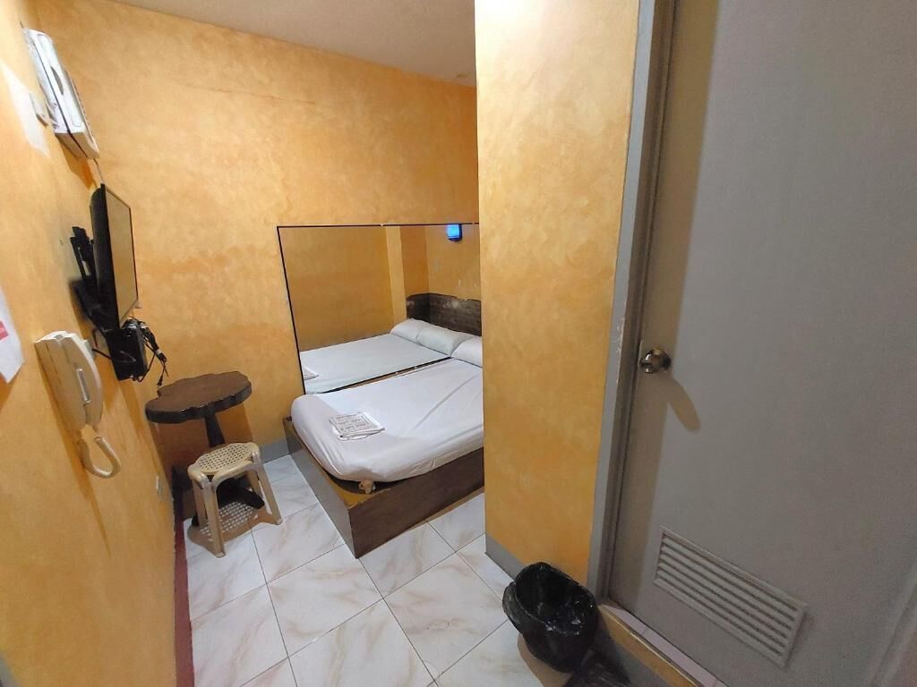 Quarto pequeno e compacto com uma cama, um banquinho, um telefone e uma lixeira.
