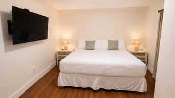 Quarto de hotel com uma cama grande, duas mesas de cabeceira com luminárias, uma TV de tela plana na parede e piso de madeira.